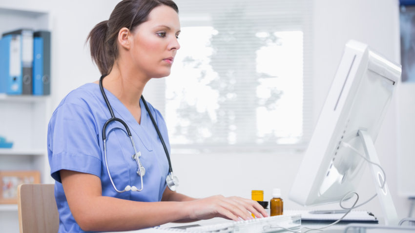 Side view of young female nurse using computer at desk in clinic ...